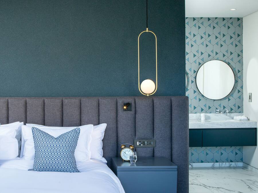 Quarto de hotel moderno com uma cama grande, duas mesas de cabeceira, um criado-mudo com relógio e banheiro com espelho e pia.
