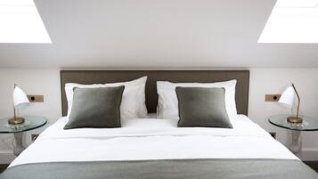 Cama bem arrumada com cabeceira cinza, duas mesas de cabeceira com luminárias e um claraboia acima.