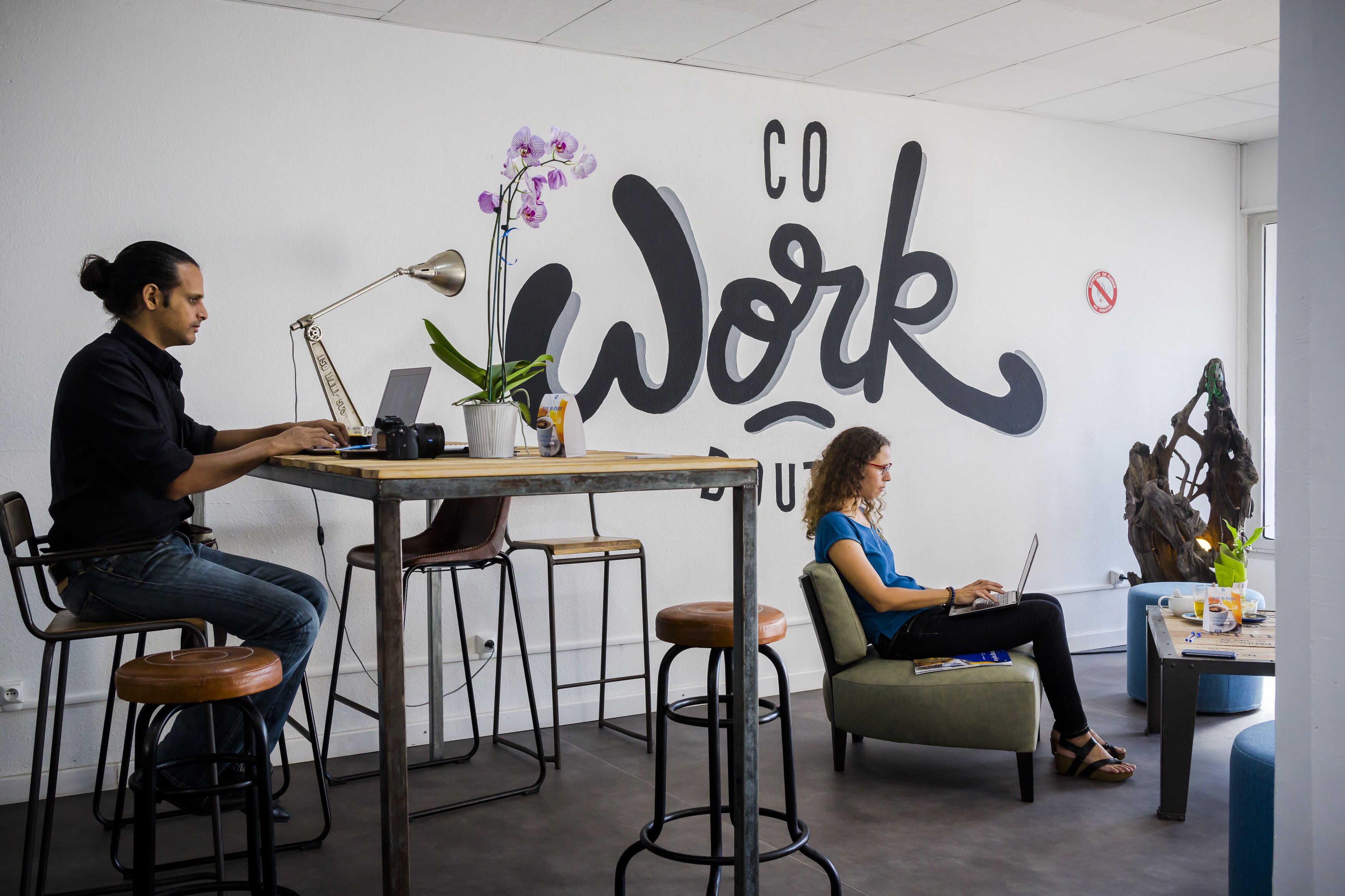 Um espaço de escritório moderno com uma grande placa na parede com a inscrição "CO Work". Duas pessoas trabalham em mesas, uma com um laptop e outra com uma câmera. O ambiente tem um design minimalista com uma mesa de madeira, banquetas e um vaso de planta.