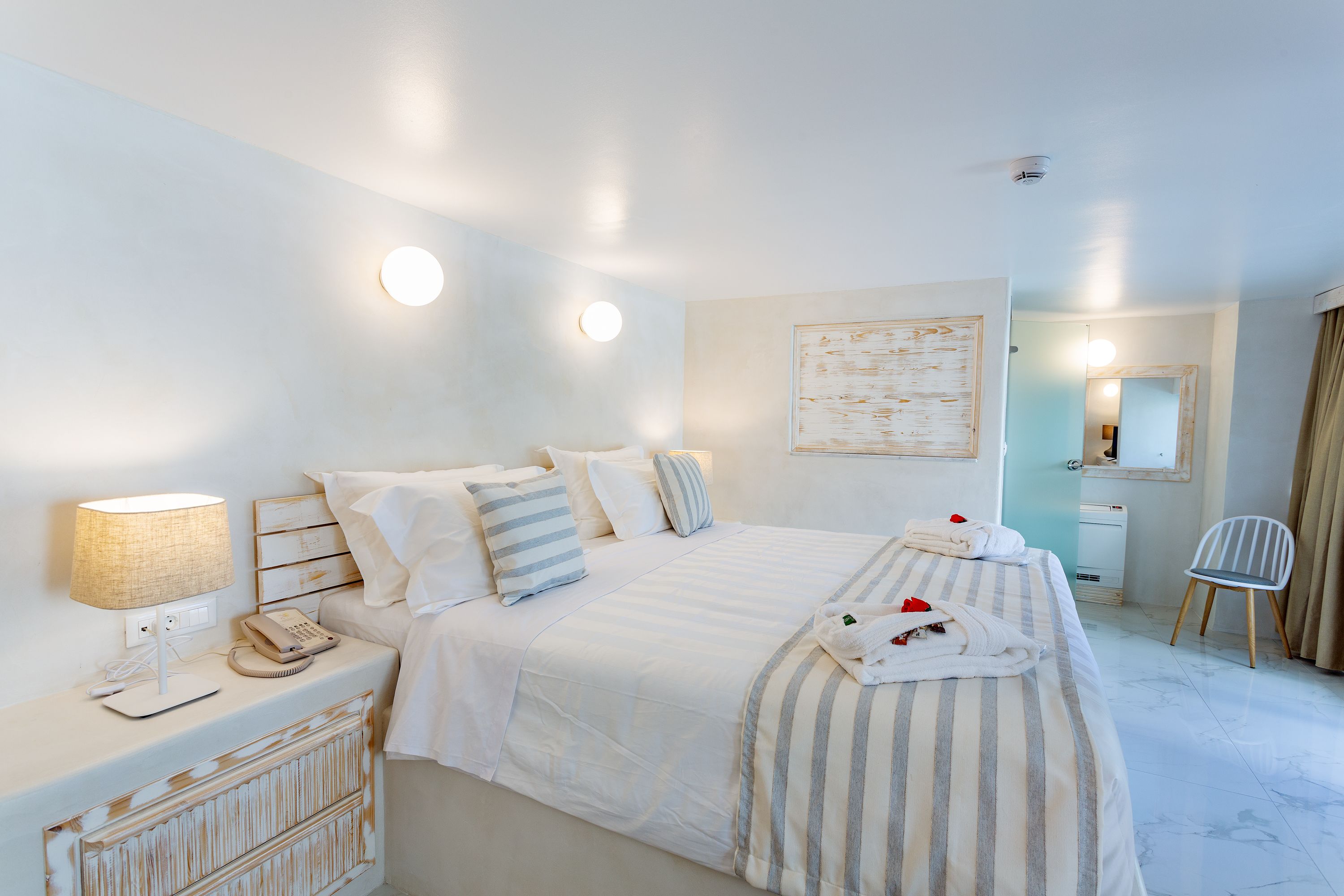 Quarto de hotel com cama bem arrumada, mesa de cabeceira com abajur, telefone e uma cadeira.