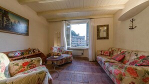 Uma sala de estar acolhedora com móveis estampados floralmente, uma janela ampla com vista para prédios e um teto com vigas de madeira aparentes.