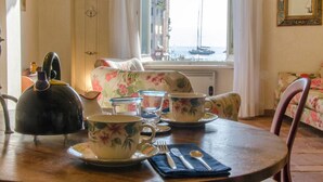 Mesa de jantar posta com xícaras e pires de padrão floral, chaleira e talheres. Uma janela com vista para edifícios e um barco é visível ao fundo.