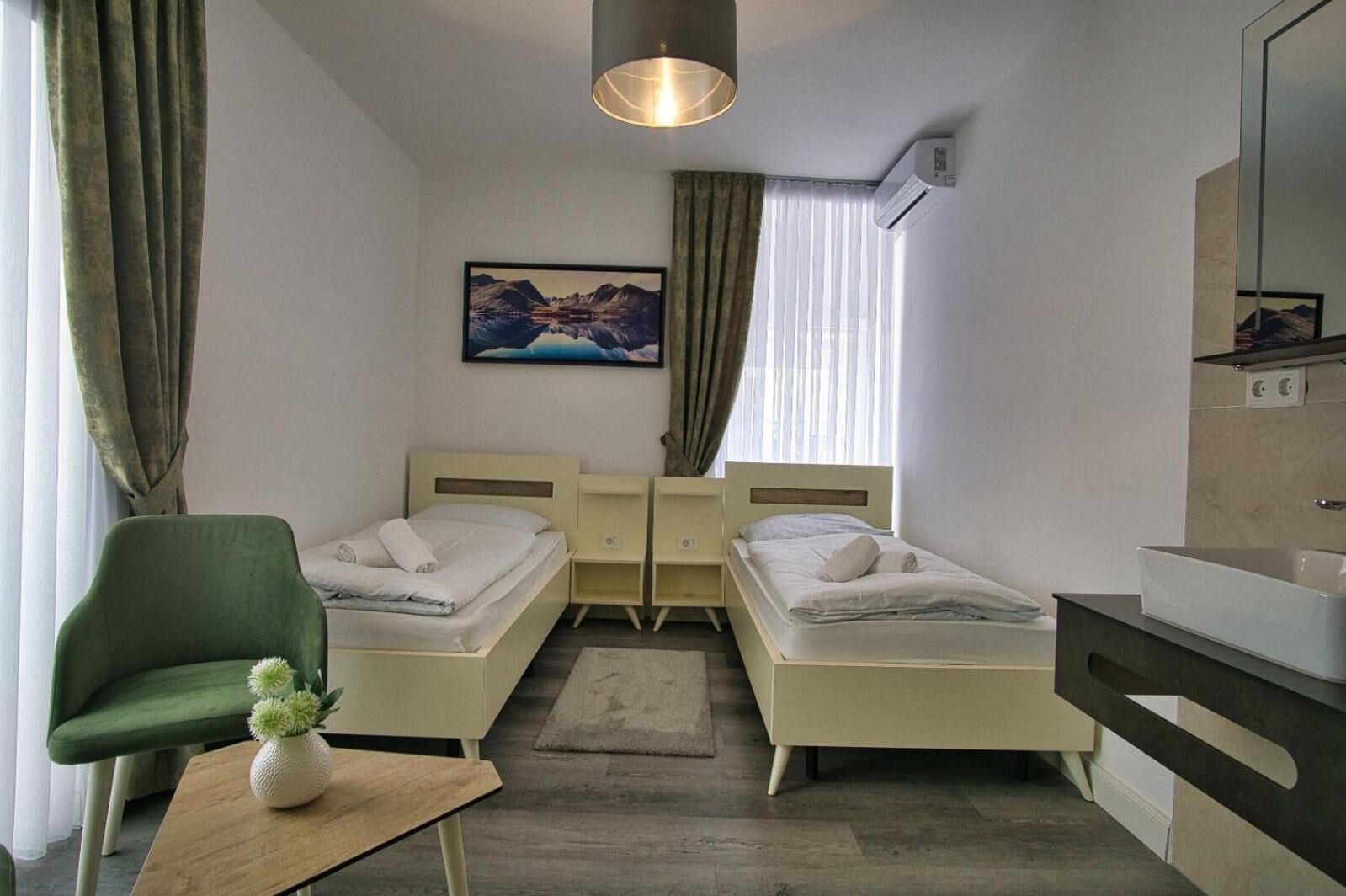 Quarto de hotel com duas camas de solteiro, uma poltrona verde, uma mesa pequena com uma planta, um quadro na parede e um banheiro com espelho e pia.