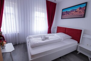 Uma cama bem arrumada com roupa de cama branca e cabeceira vermelha, uma mesa de cabeceira branca com um vaso de flores e um quadro de uma paisagem montanhosa na parede.
