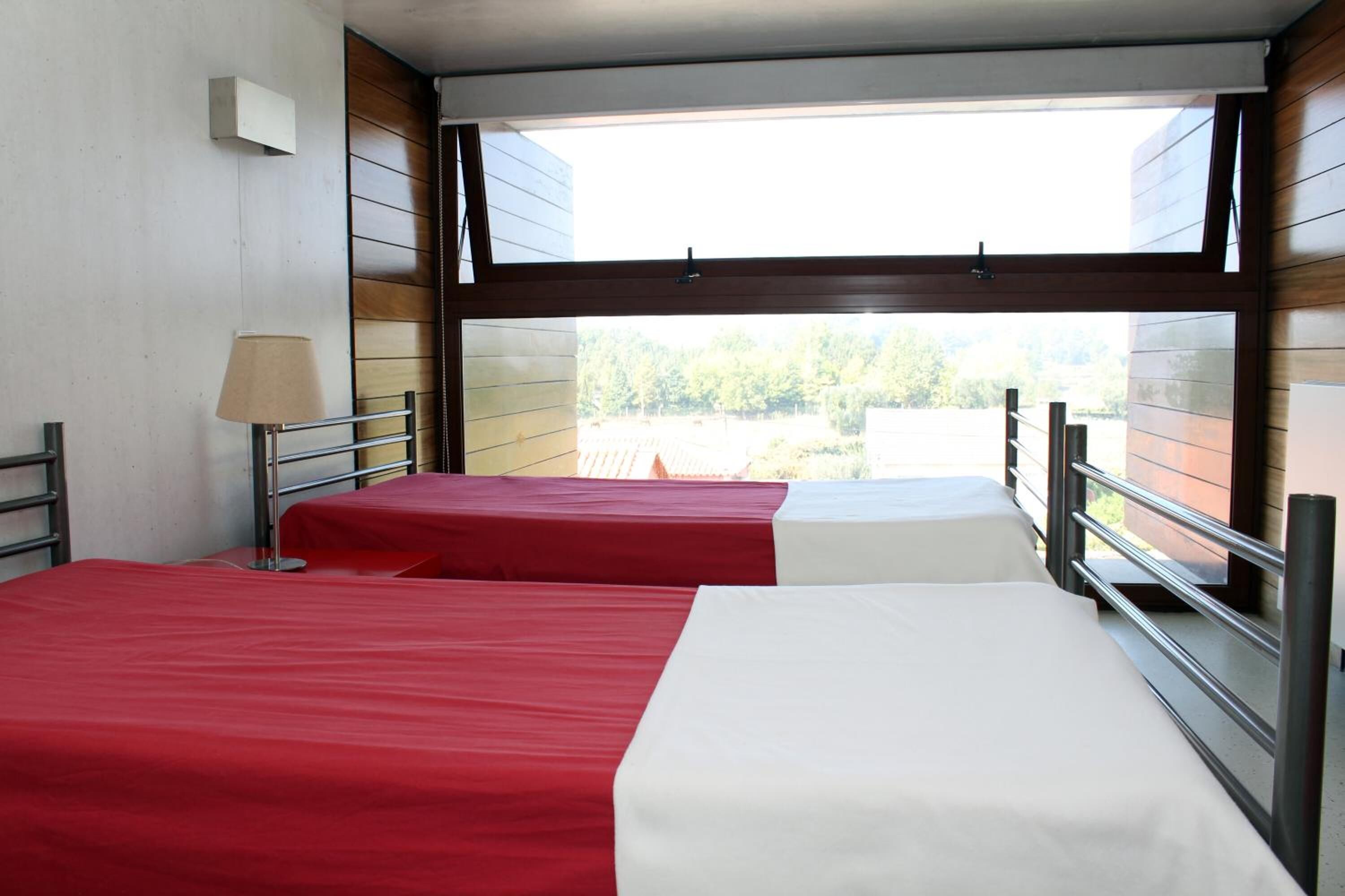Quarto com duas camas, uma com colcha vermelha e outra com colcha branca, uma luminária e janela com vista para árvores e edifícios.