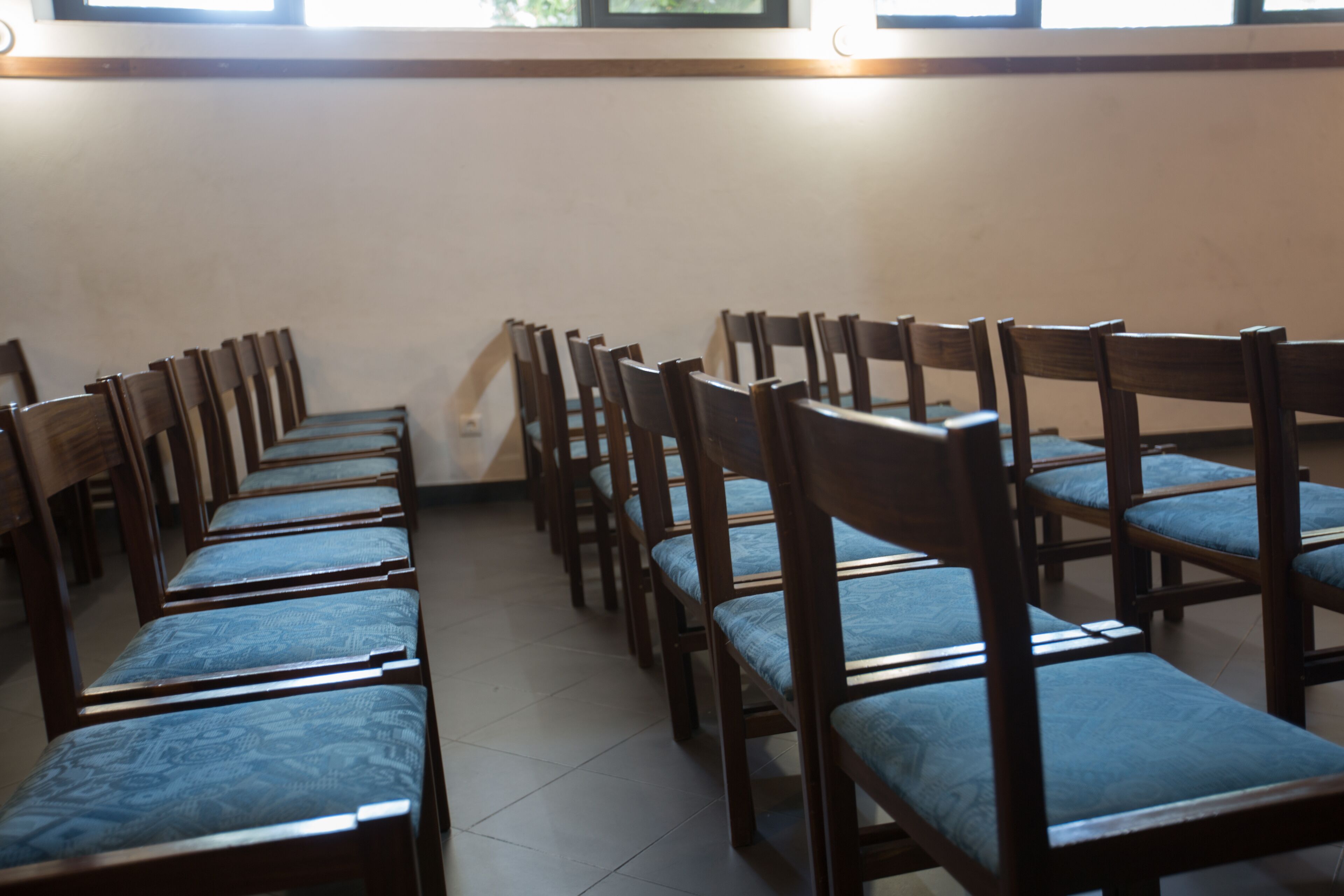 Fileiras de cadeiras de madeira com almofadas azuis dispostas em um ambiente de sala de aula ou reunião.