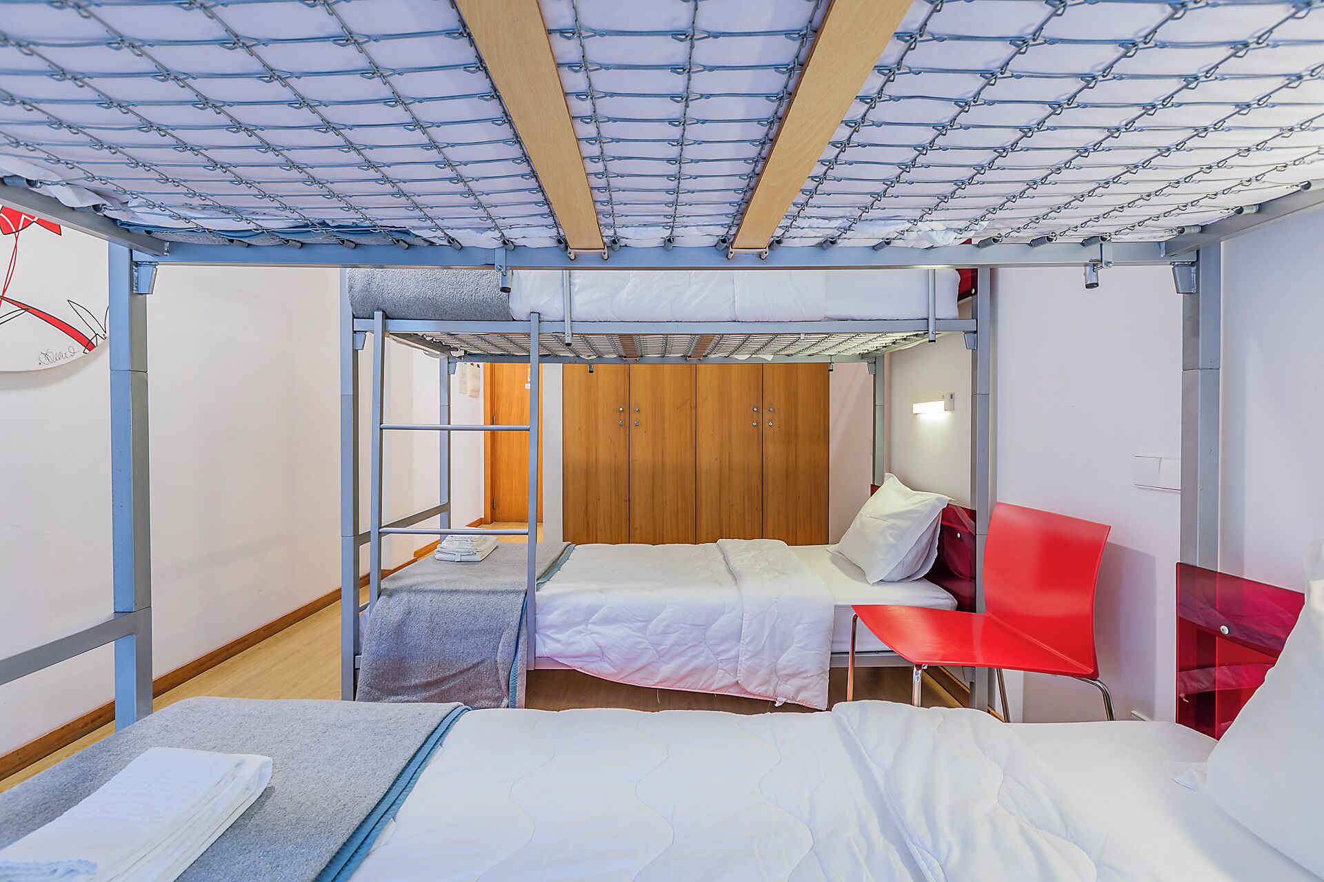 Quarto pequeno com beliches, uma cadeira vermelha e um guarda-roupa de madeira.