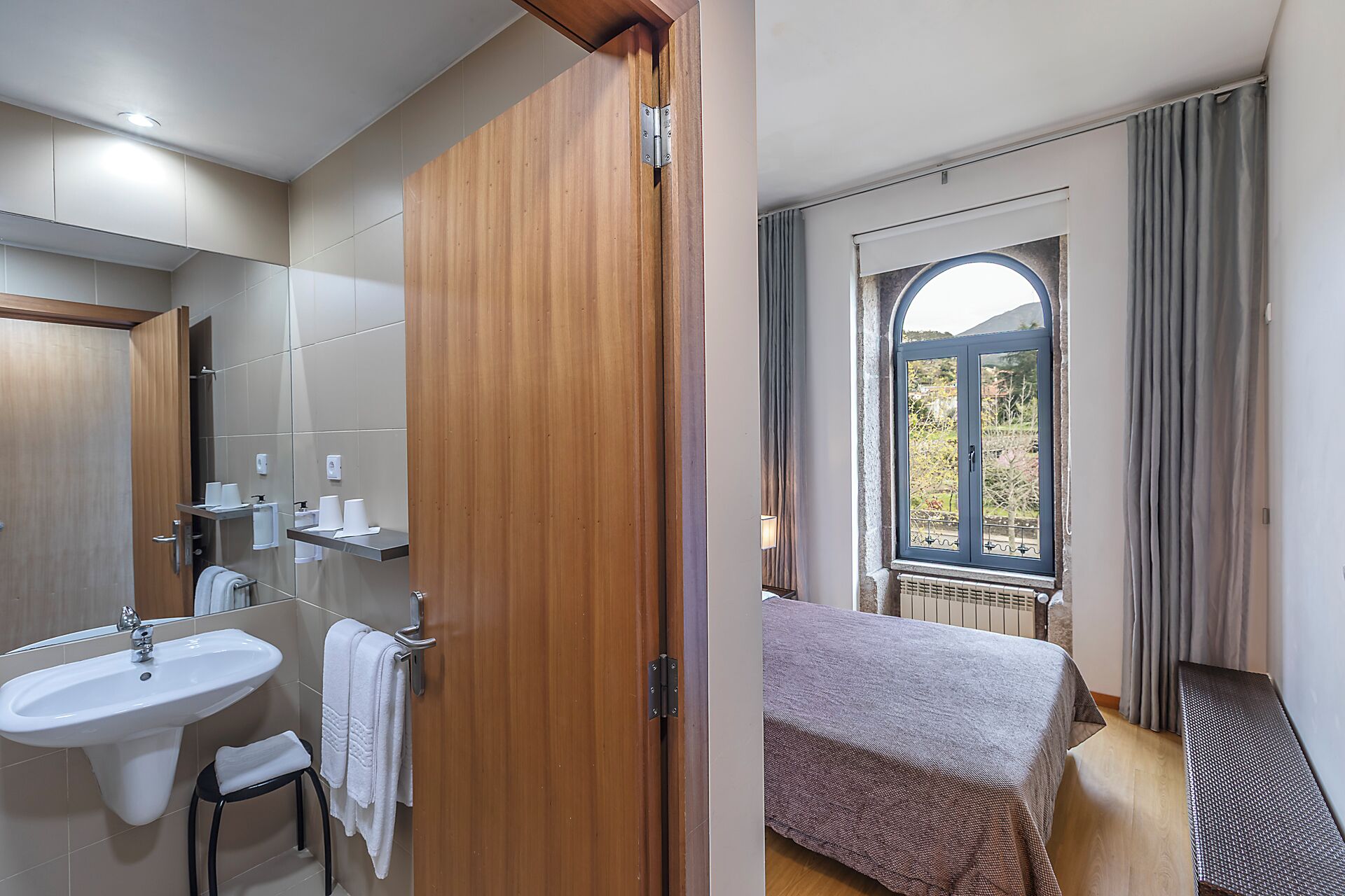 Quarto de hotel com porta de madeira, pia, espelho, cama e janela com vista.