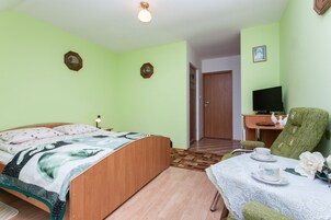 Quarto com cama, mesa com toalha e chávenas, poltrona verde, televisão sobre uma secretária de madeira e dois relógios fixados na parede.