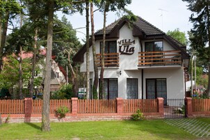 Uma villa Ula de dois andares com varanda, cercada por um muro de tijolos e árvores.