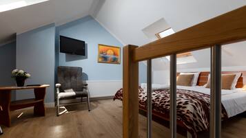 Quarto moderno com teto inclinado, TV montada, um quadro, colcha de cama com estampa de leopardo e uma cadeira de madeira.
