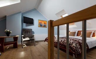 Quarto moderno com teto inclinado, TV montada, um quadro, colcha de cama com estampa de leopardo e uma cadeira de madeira.