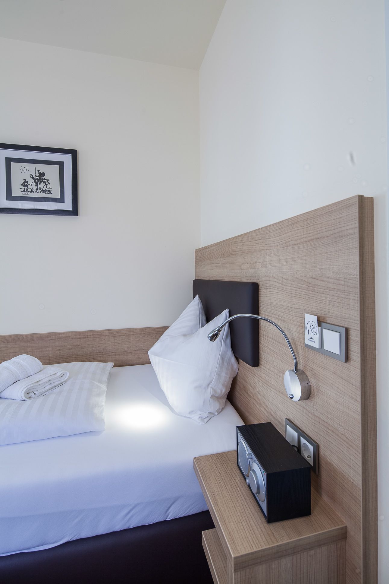 Quarto de hotel com uma cama bem arrumada, uma mesa de cabeceira com um rádio, uma luminária na parede e um quadro na parede.