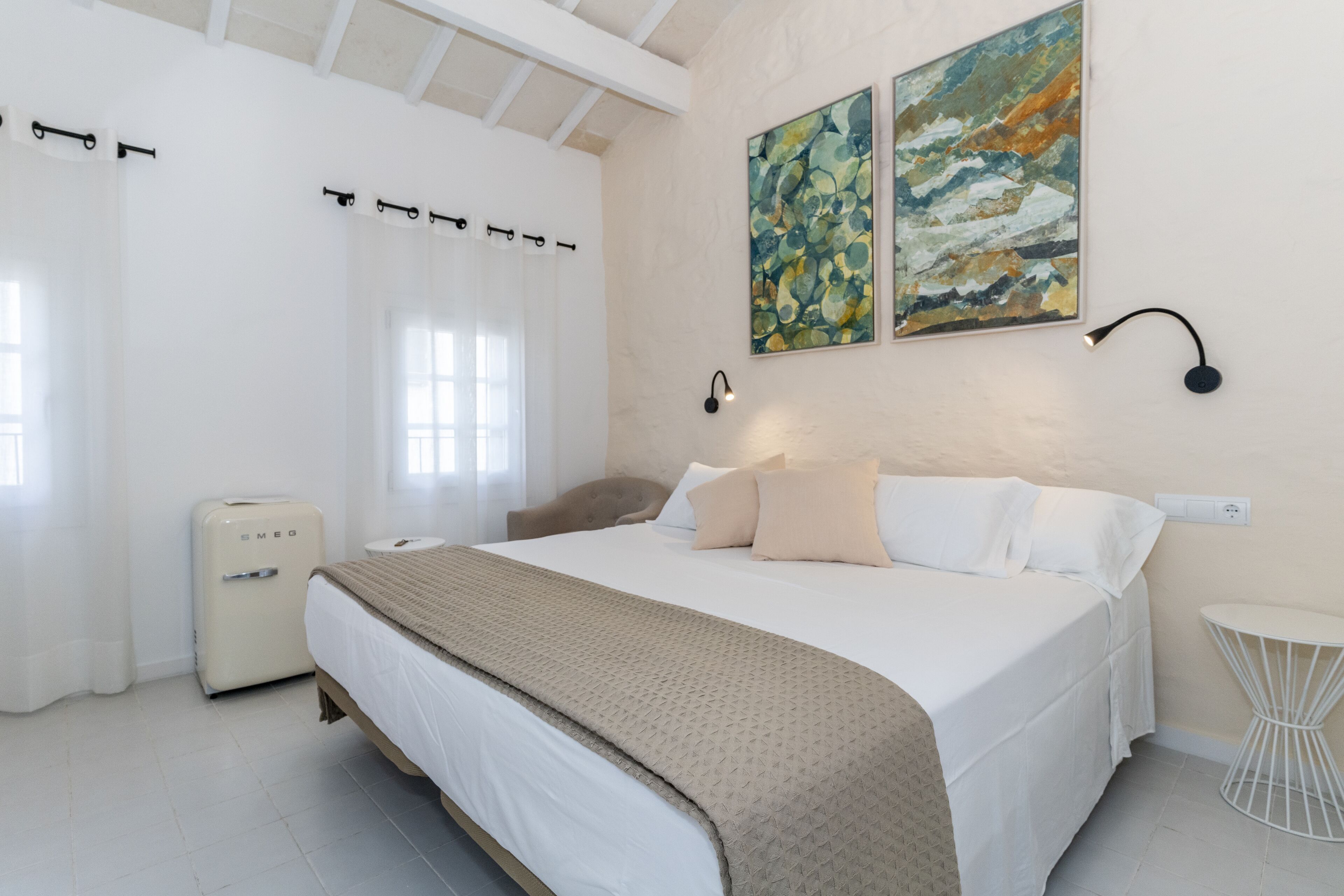 Quarto com cama, cortinas, luminária na parede e uma pequena mesa.