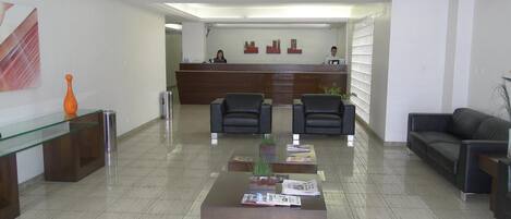 Um hall amplo com um balcão de recepção, dois sofás de couro preto, uma mesa de centro de vidro com revistas e um vaso laranja decorativo.