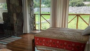 Quarto com cama, parede de pedra e vista para uma área gramada por grandes janelas.