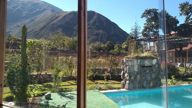 Área de piscina com montanhas ao fundo, rodeada por um jardim com diversas plantas e uma estrutura de pedra.