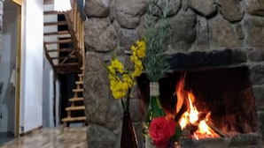 Uma lareira de pedra com fogo, uma escadaria de madeira e um vaso com flores sobre uma mesa.