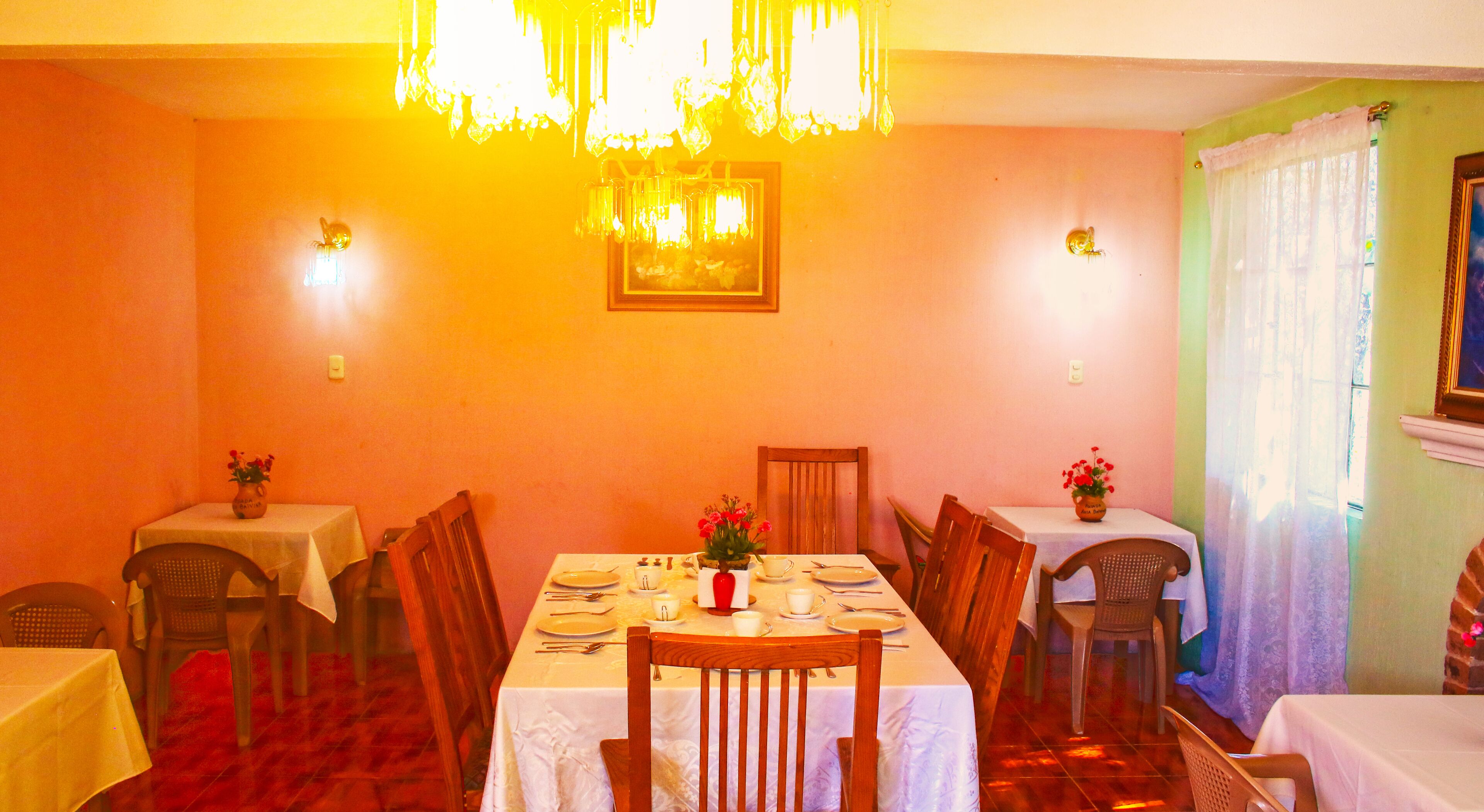 Uma sala de jantar com uma mesa comprida posta para quatro pessoas, rodeada por cadeiras de madeira. Há também duas mesas menores com cadeiras adicionais, e um lustre pendurado no teto.