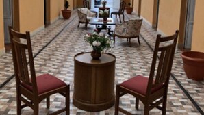 Um corredor com piso de azulejos, cadeiras de madeira com almofadas vermelhas, uma mesa redonda central com um vaso de flores e teto com claraboia.