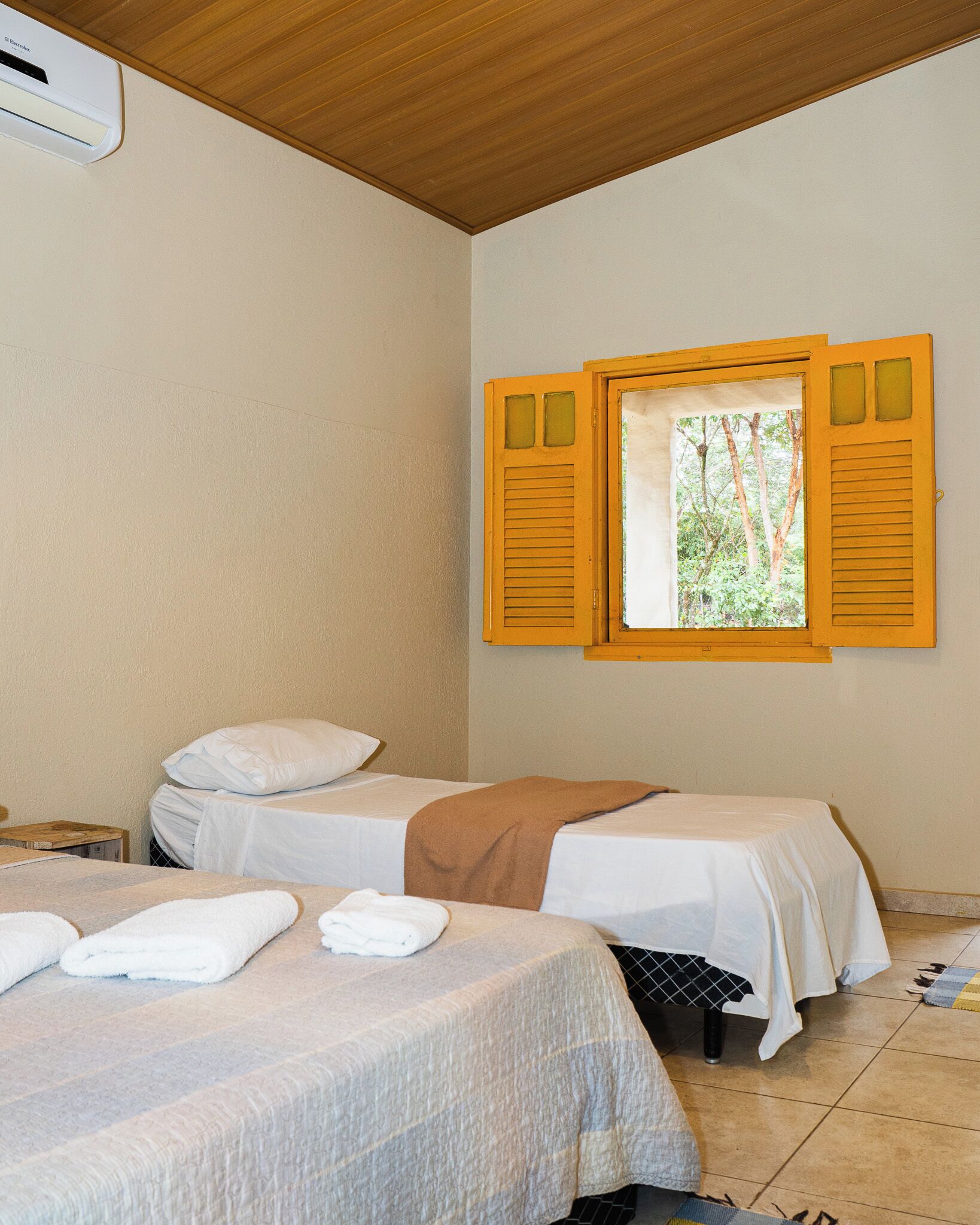 Quarto com duas camas, janela com persianas, ventilador de teto e ar condicionado fixado na parede.