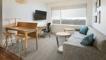 Uma sala de estar moderna com uma escrivaninha de madeira, dois bancos altos, um sofá cinza com almofadas azuis, uma mesa de centro redonda e uma janela grande com vista para a cidade.