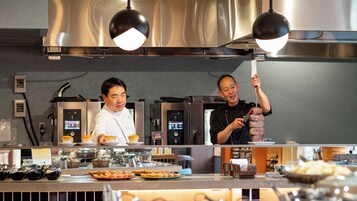 Dois chefs em cozinha equipada com eletrodomésticos de aço inoxidável e uma variedade de alimentos.