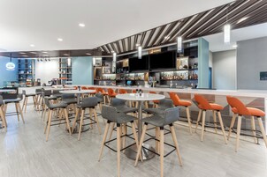 Uma área de bar moderna com mesas e cadeiras redondas, um balcão com visor digital e prateleiras repletas de garrafas.