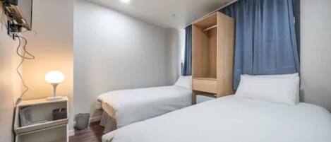 Quarto de hotel com duas camas, um guarda-roupa, uma mesa de cabeceira com abajur e uma televisão na parede.