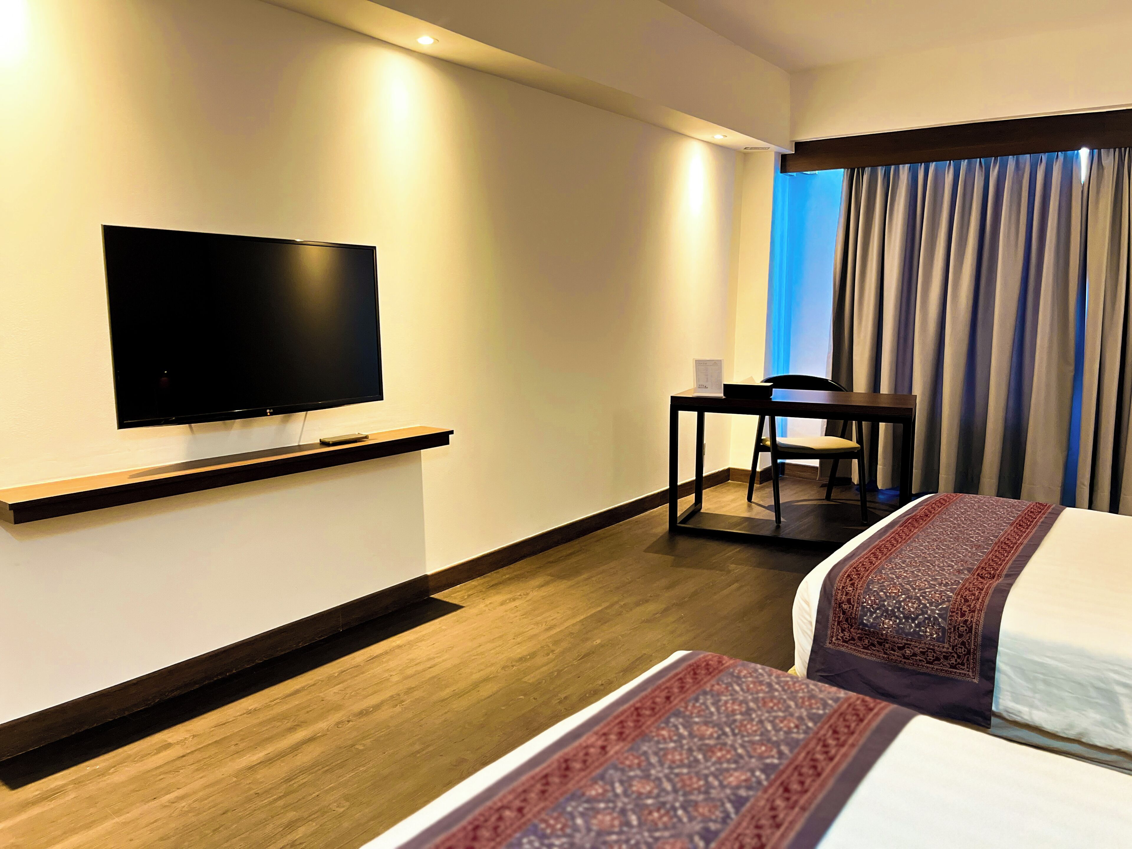 Quarto de hotel com televisão de tela plana na parede, uma escrivaninha com cadeira e uma cama com colcha estampada.