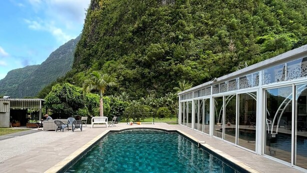 Uma piscina com borda de pedra, rodeada por um pátio com cadeiras e um revestimento de vidro.