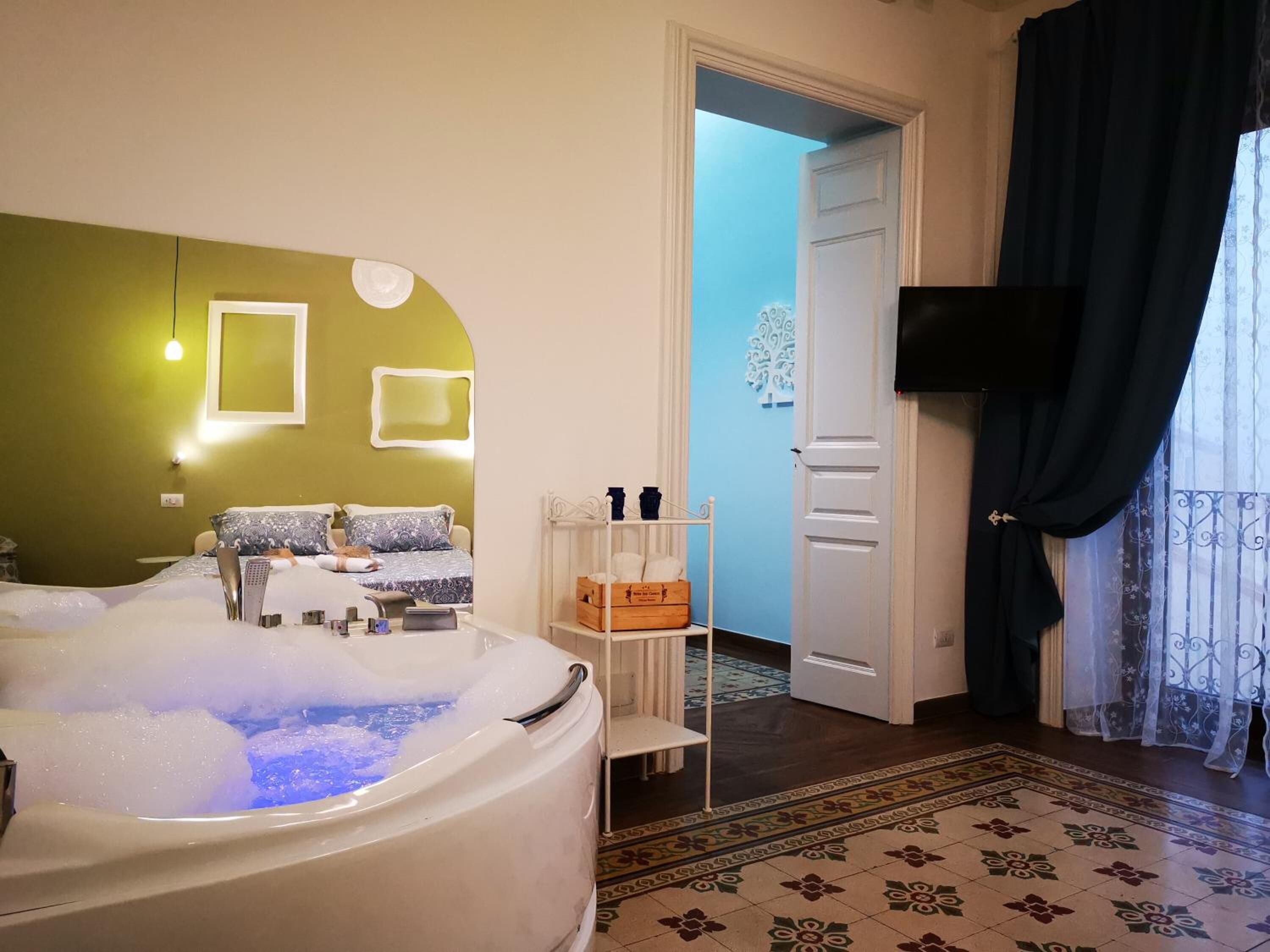 Quarto com hidromassagem, cama com roupa de cama estampada, prateleira branca pequena com objetos, porta que dá para outro cômodo e piso estampado.