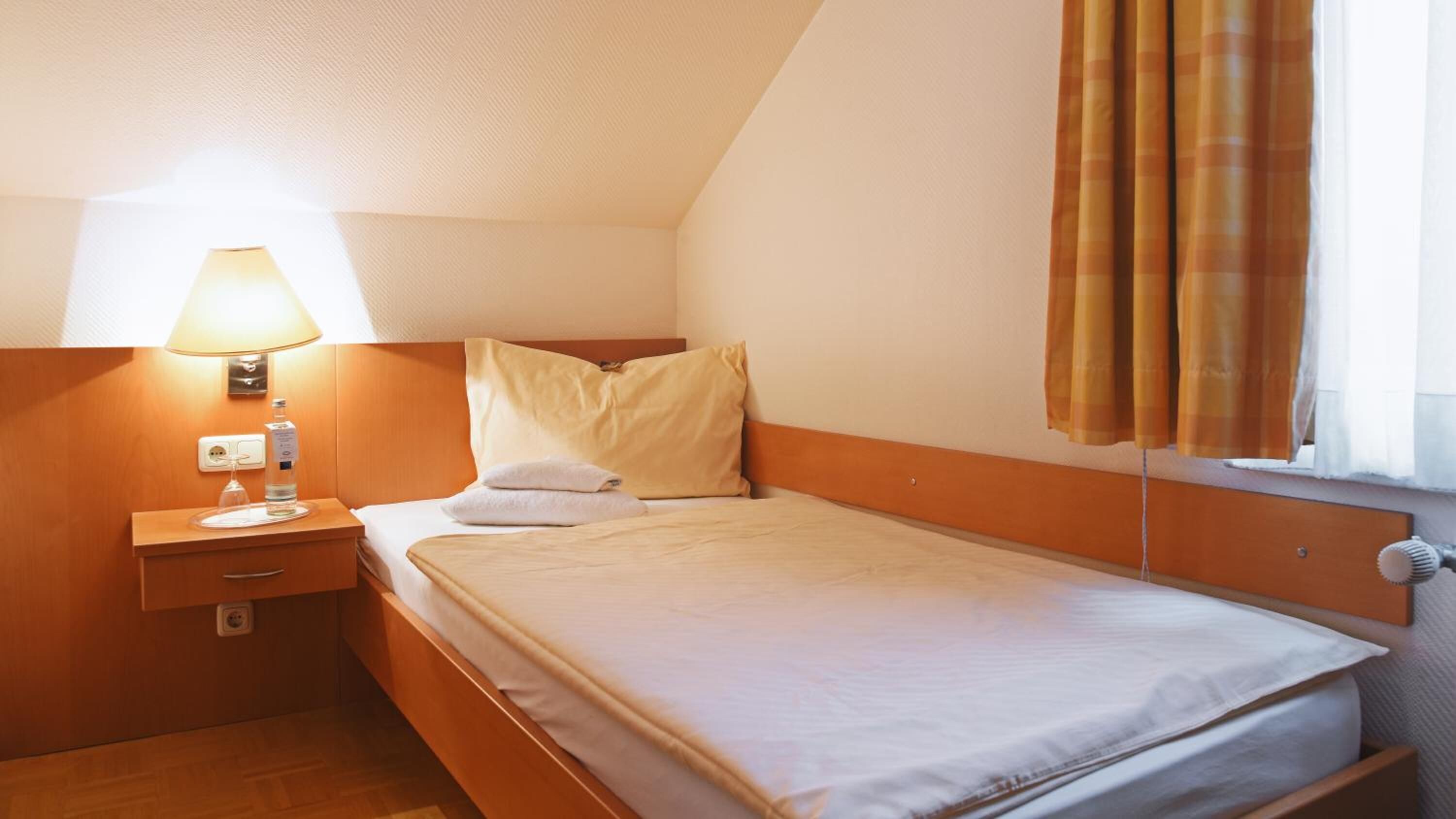 Uma cama de solteiro em quarto com cabeceira de madeira, mesa de cabeceira com abajur e janela com cortinas.