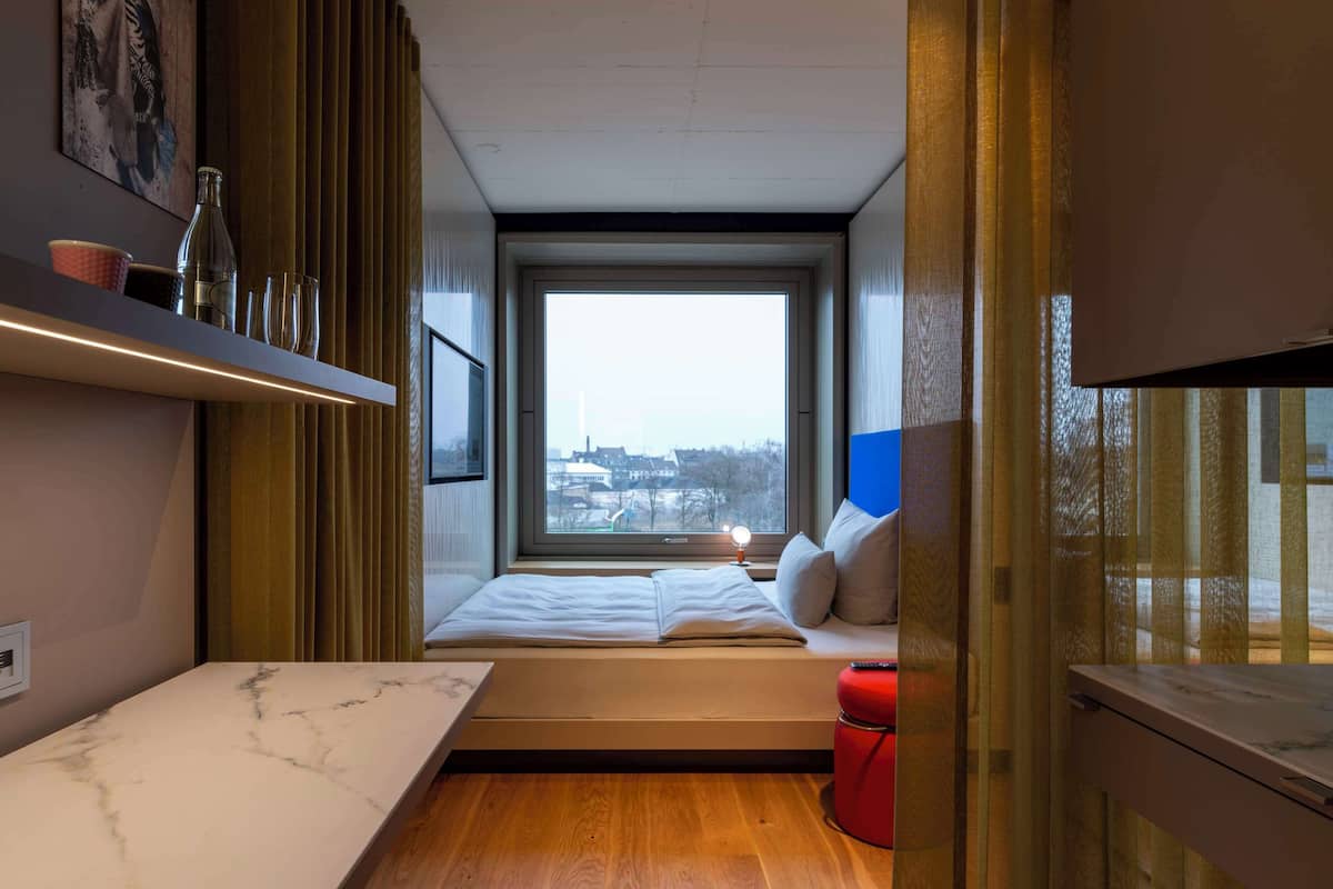 Quarto compacto com cama, uma mesinha e janela com vista para a cidade.