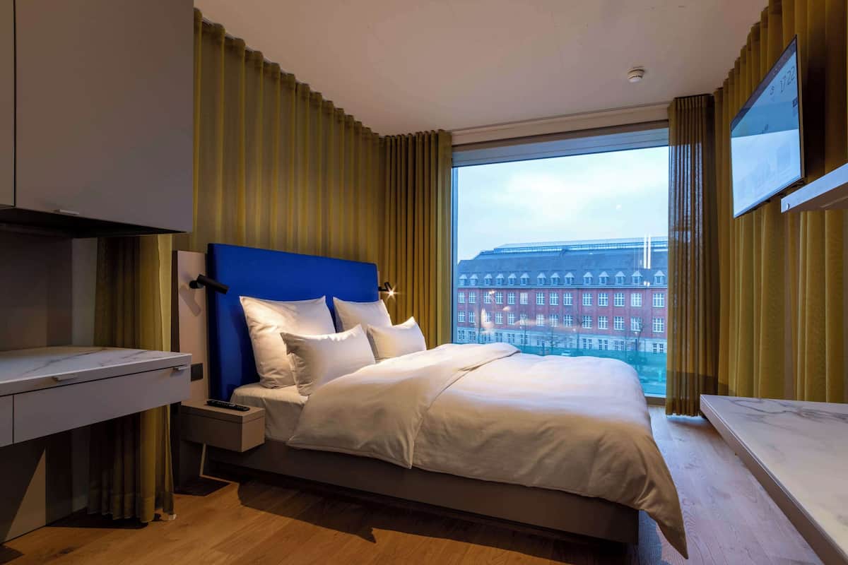 Quarto de hotel moderno com uma cama grande, uma escrivaninha e vista para a cidade através de uma janela ampla.