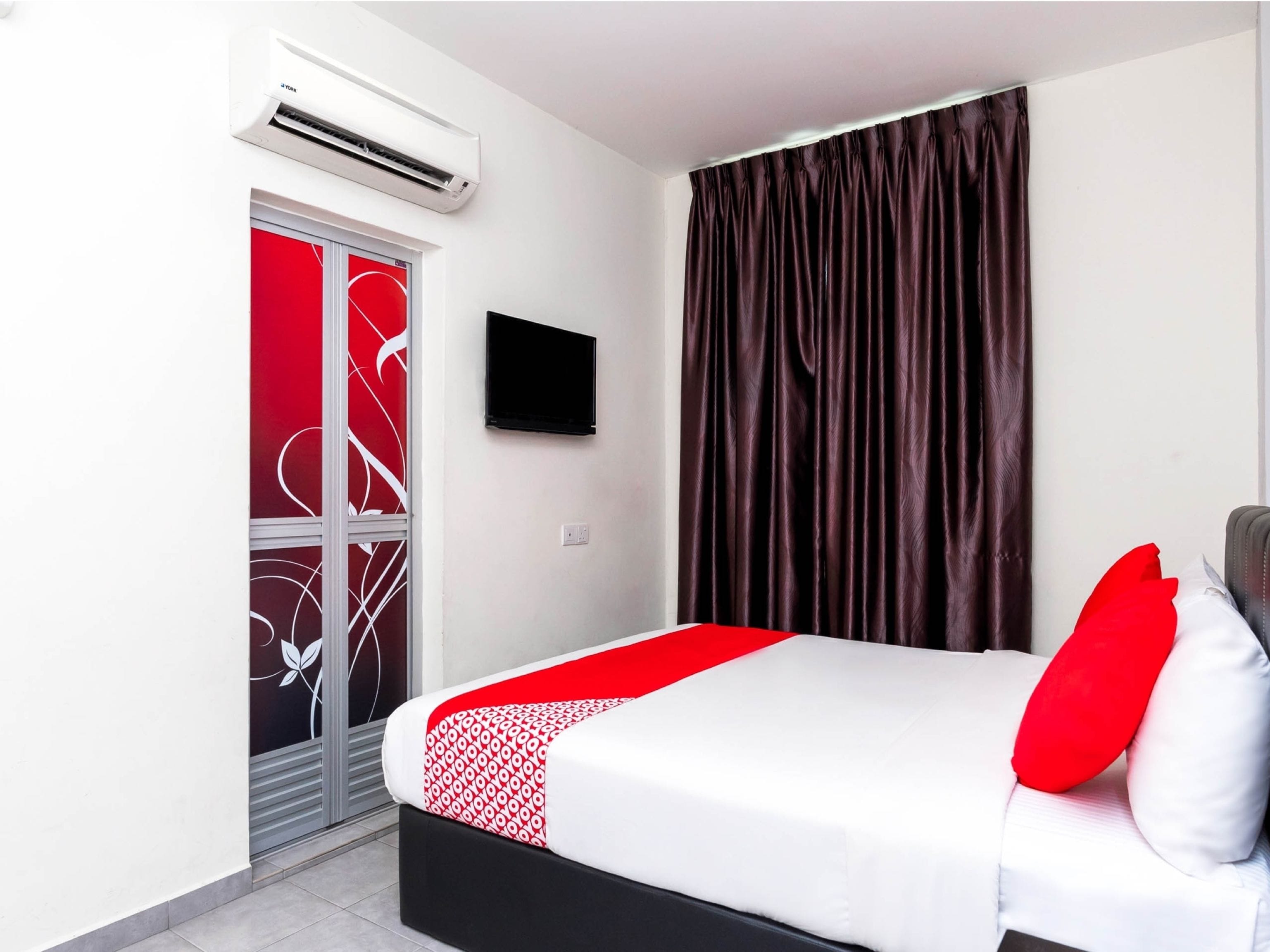 Quarto de hotel com cama, televisão, ar condicionado, janela com cortinas e porta com painel de vidro decorativo.