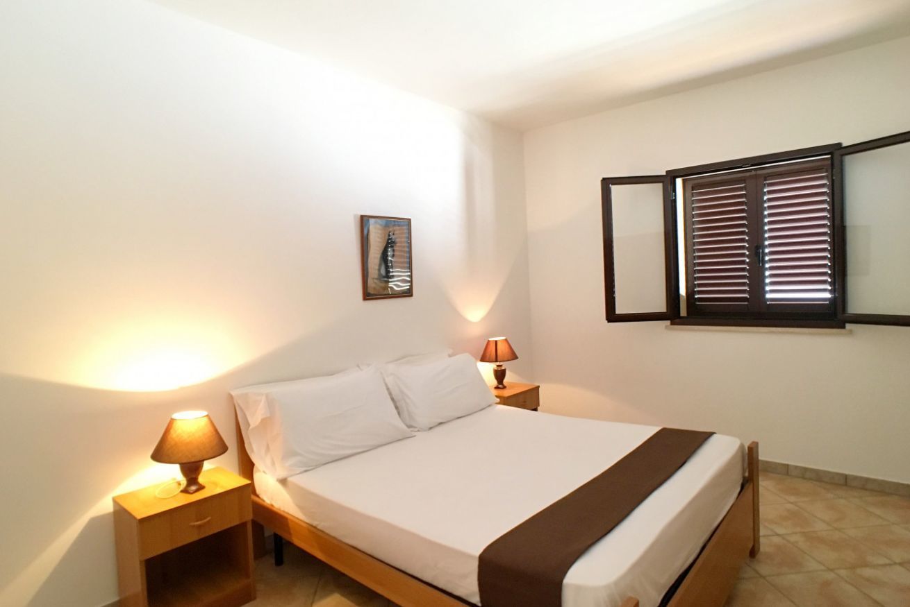 Uma cama de solteiro com estrutura de madeira, roupa de cama branca e um cobertor marrom. Há duas mesas de cabeceira com luminárias, uma de cada lado da cama. Um quadro emoldurado está pendurado na parede acima da cama.