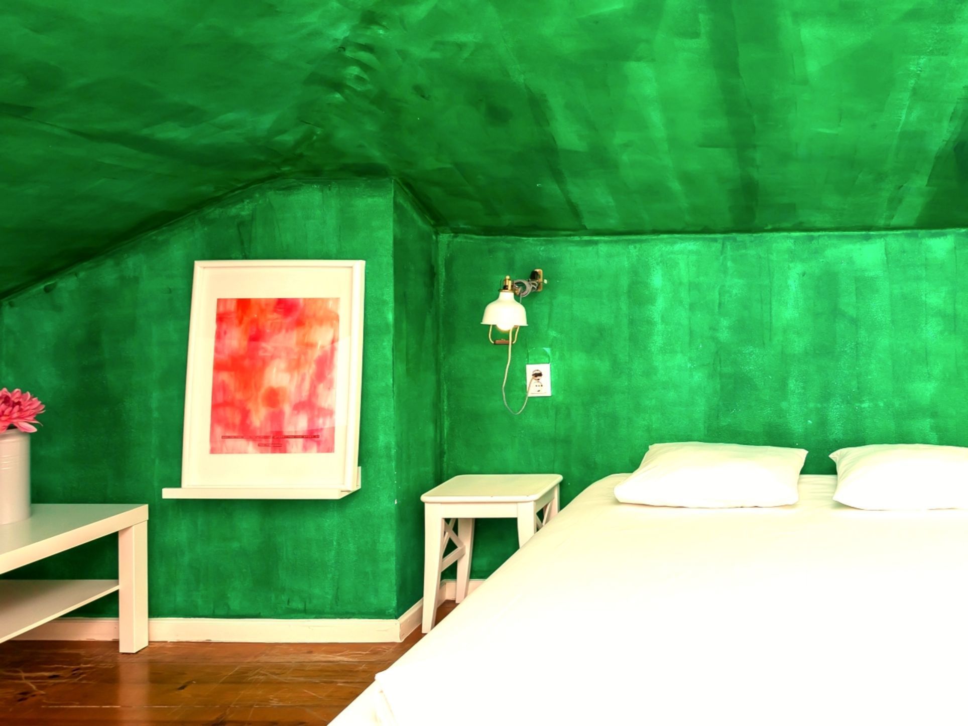 Quarto com paredes verdes, cama branca, mesinha de cabeceira pequena, um criado-mudo e um quadro emoldurado, com uma luminária fixada na parede.