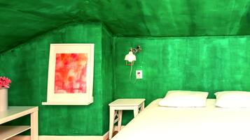 Quarto com paredes verdes, cama branca, mesinha de cabeceira pequena, um criado-mudo e um quadro emoldurado, com uma luminária fixada na parede.