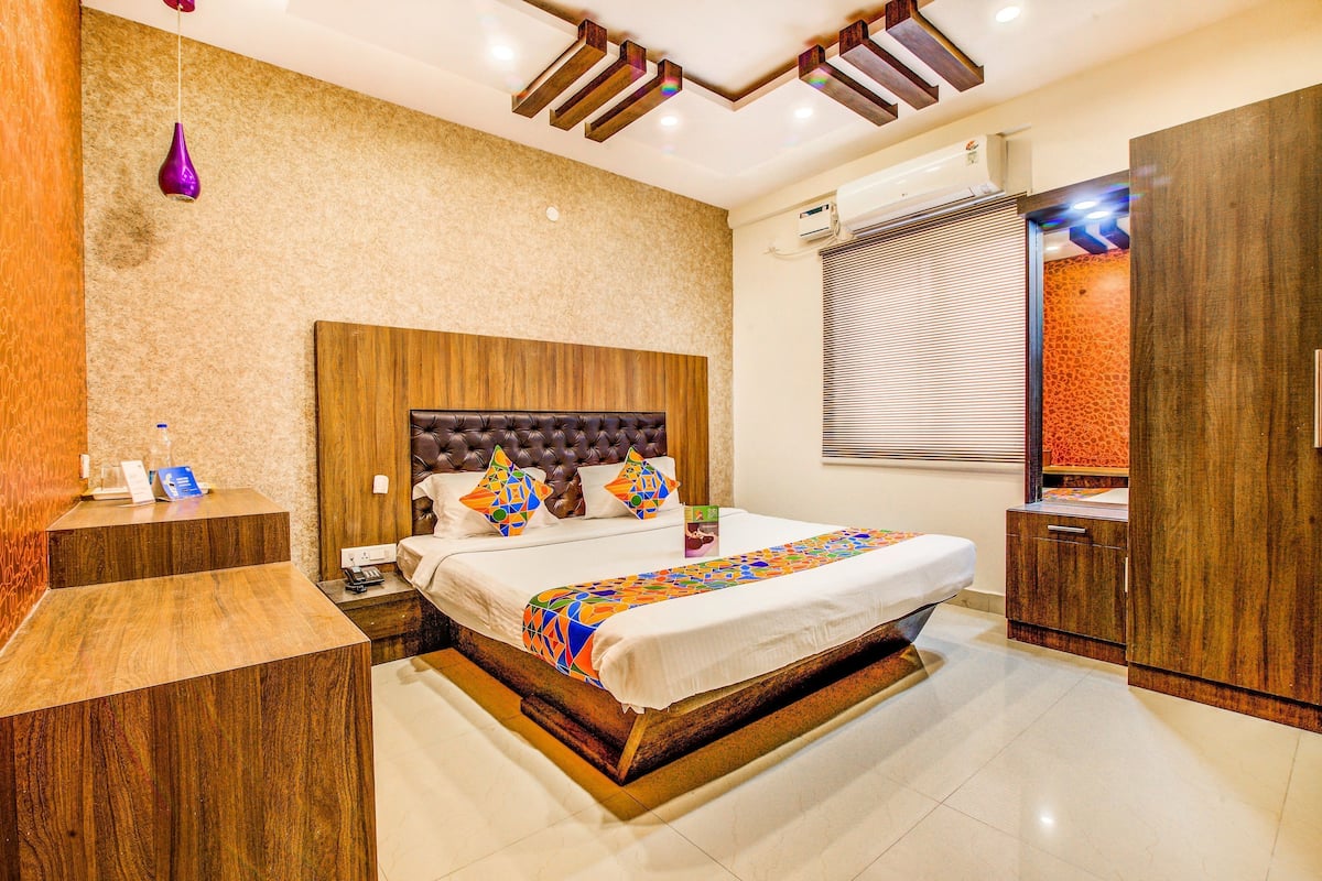 Quarto de hotel com uma cama grande, cabeceira de madeira, mesa de cabeceira, guarda-roupa e janela com persianas.