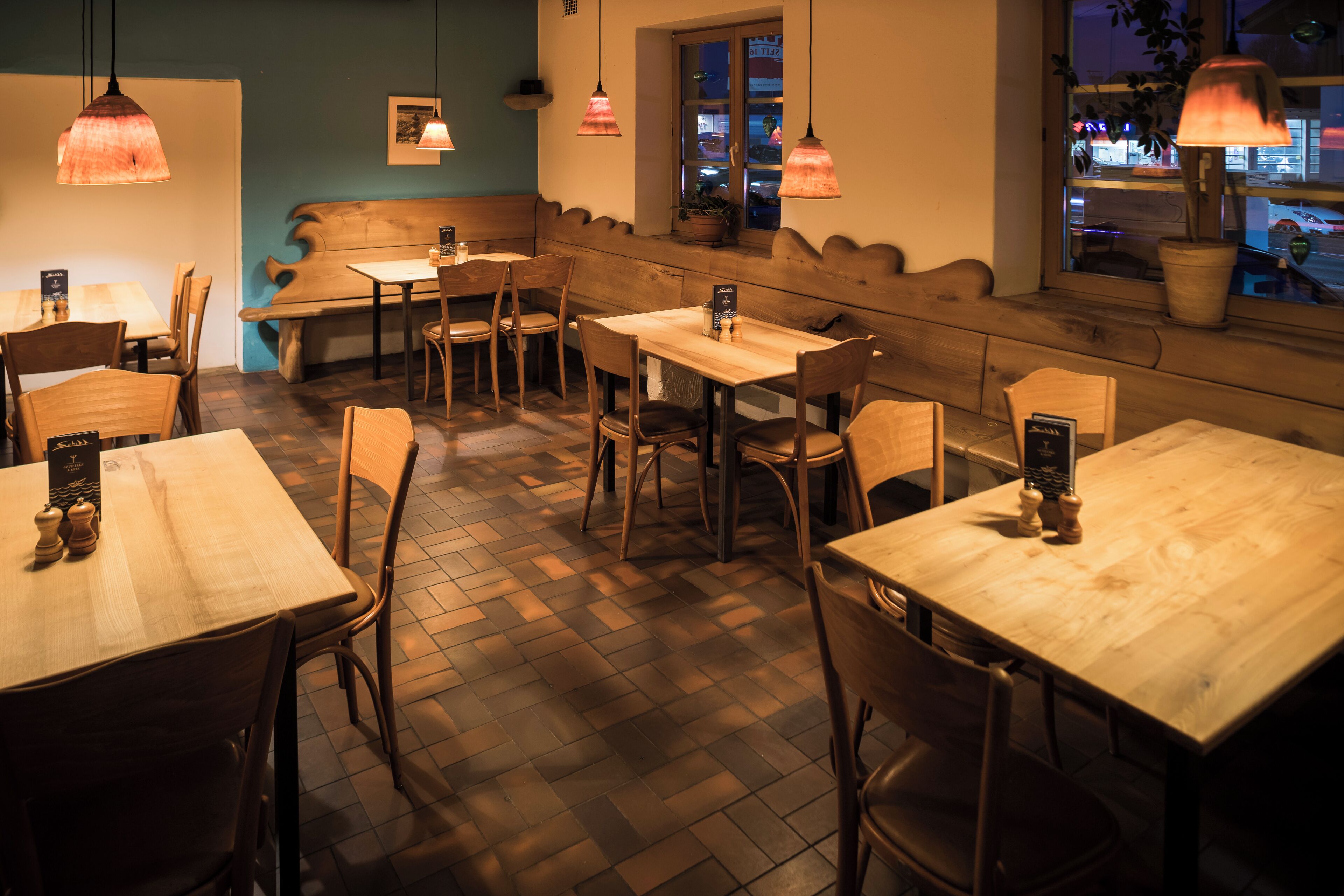 Um restaurante com mesas e cadeiras de madeira, um banco e luminárias pendentes.