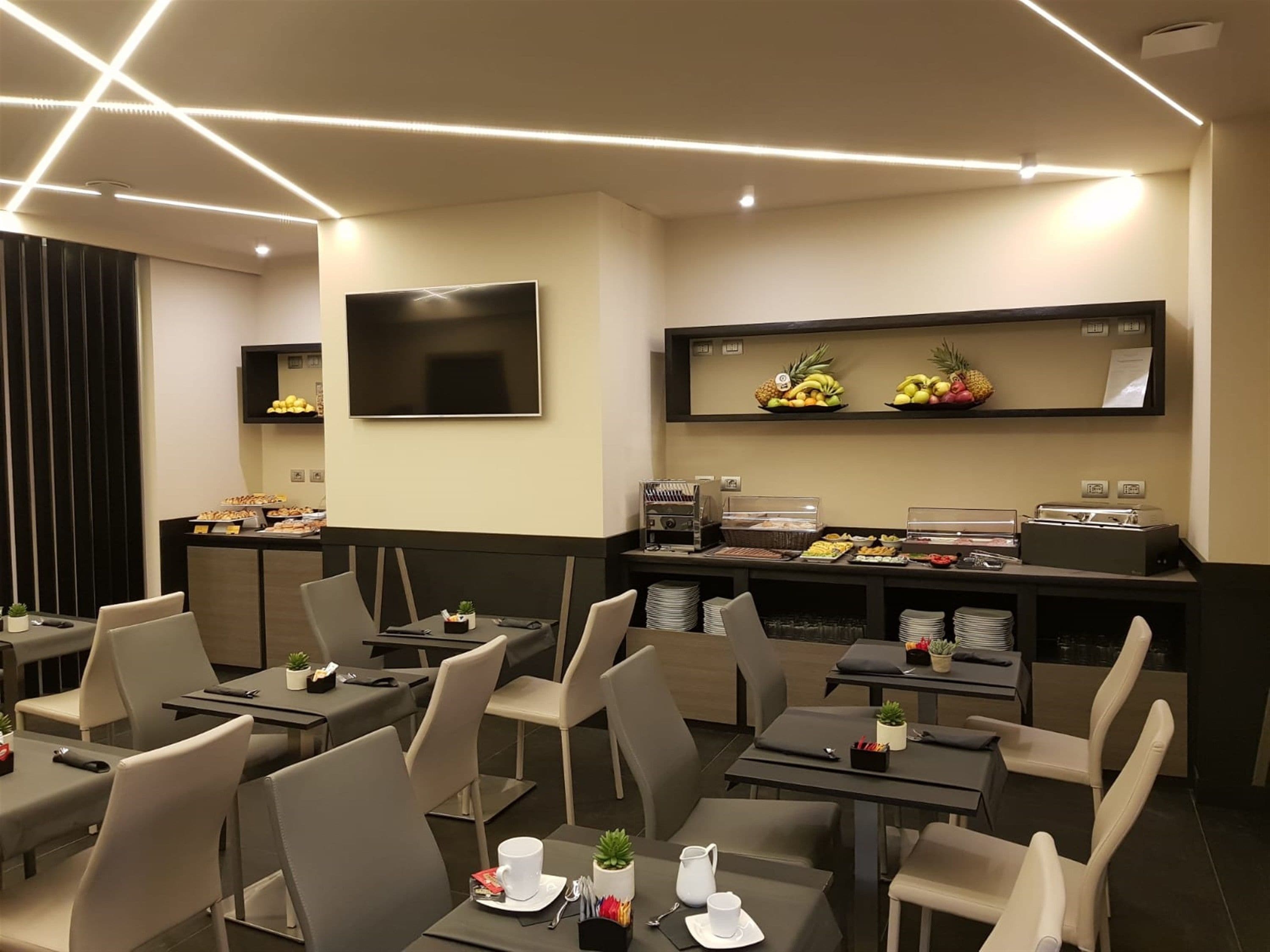 Uma área de refeições moderna com mesas e cadeiras, um buffet com frutas e bebidas, e uma TV de tela plana na parede.