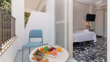 Quarto de hotel com cama, TV fixada na parede, mesinha com um cesto de frutas, um livro e uma xícara.