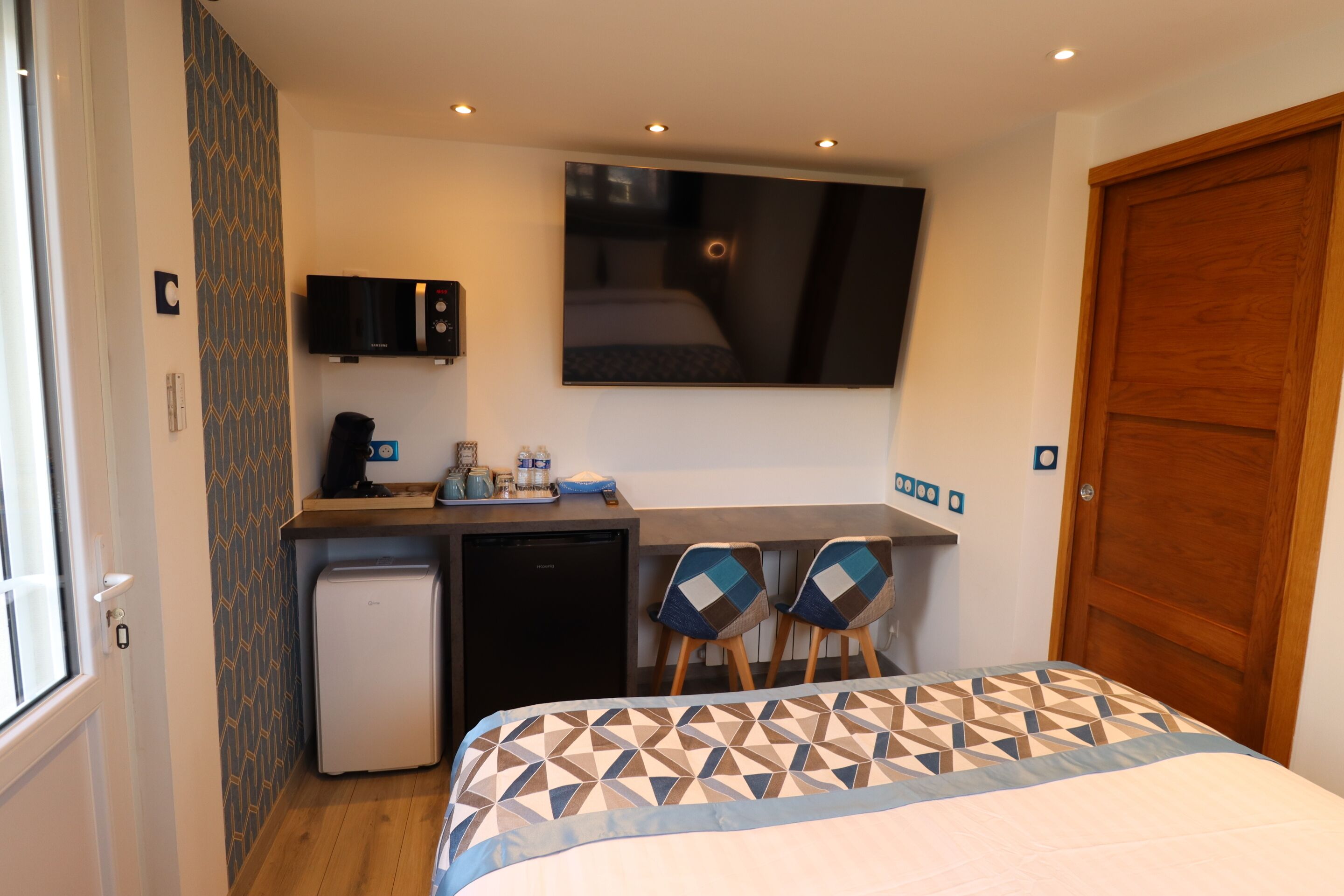 Quarto de hotel compacto com uma cama, pequeno espaço de cozinha, televisão e porta de madeira.