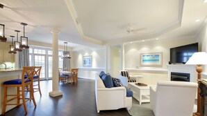 Uma sala de estar ampla com um sofá branco, uma área de refeições com cadeiras de madeira, uma lareira e uma TV grande fixada na parede.