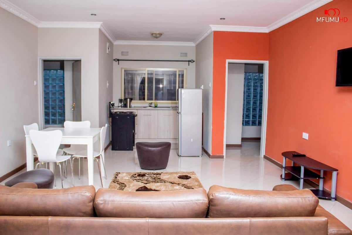 Uma sala de estar moderna com um sofá de couro marrom, uma área de jantar com cadeiras brancas, uma cozinha com geladeira e uma parede laranja.