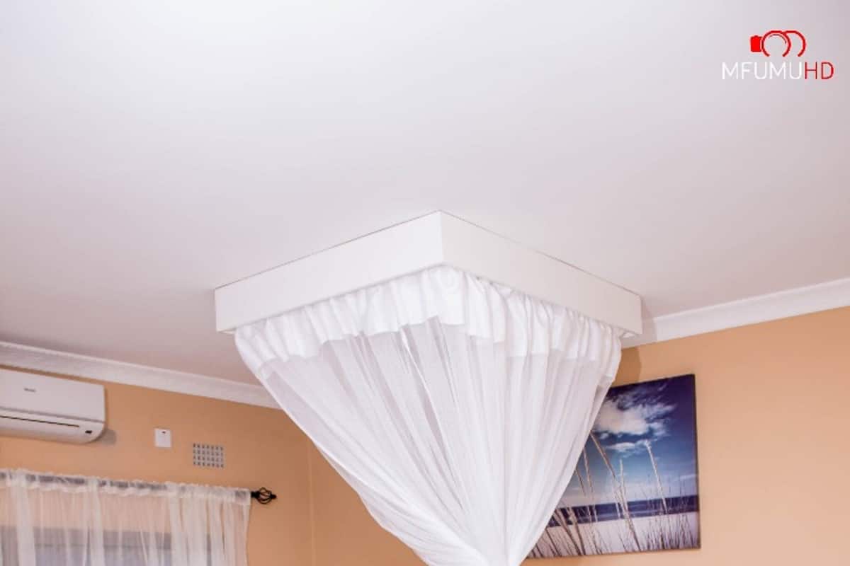 Quarto com cama, cortinas brancas, ventilador de teto, mesa de cabeceira e ar condicionado fixado na parede.