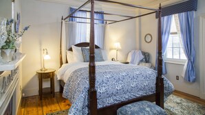 Uma cama de dossel com colcha florida, mesas de cabeceira com abajures, um espelho na parede e uma janela com cortinas.