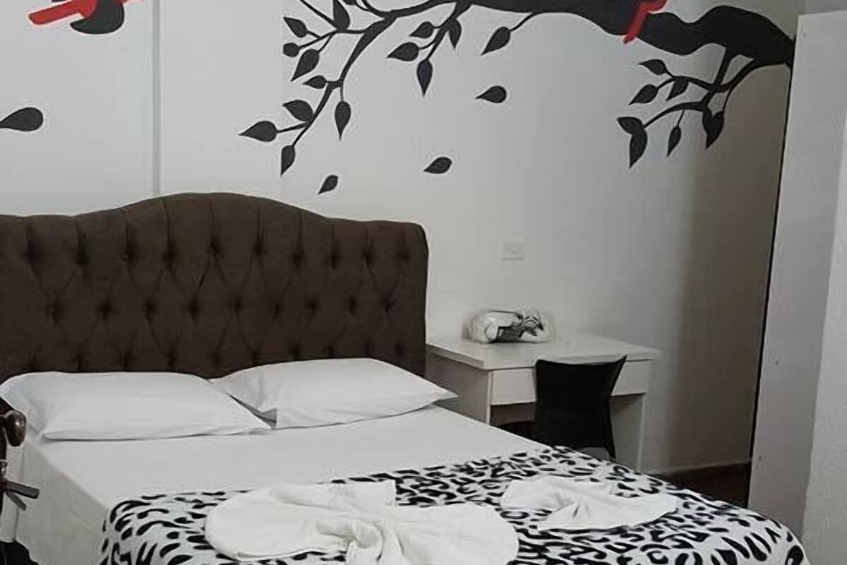 Um quarto com cabeceira estofada, cama com colcha de padrão de leopardo, mesa de cabeceira pequena e parede com adesivo de árvore e pássaros.