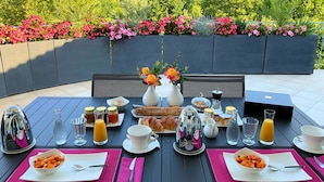 Uma área de refeições ao ar livre com uma mesa posta para o café da manhã, incluindo croissants, frutas e bebidas.